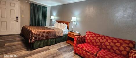 Quarto de hotel com cama, mesa de cabeceira, sofá vermelho estampado e uma porta.