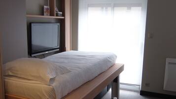 Quarto com cama, televisão presa à parede e janela com persianas.