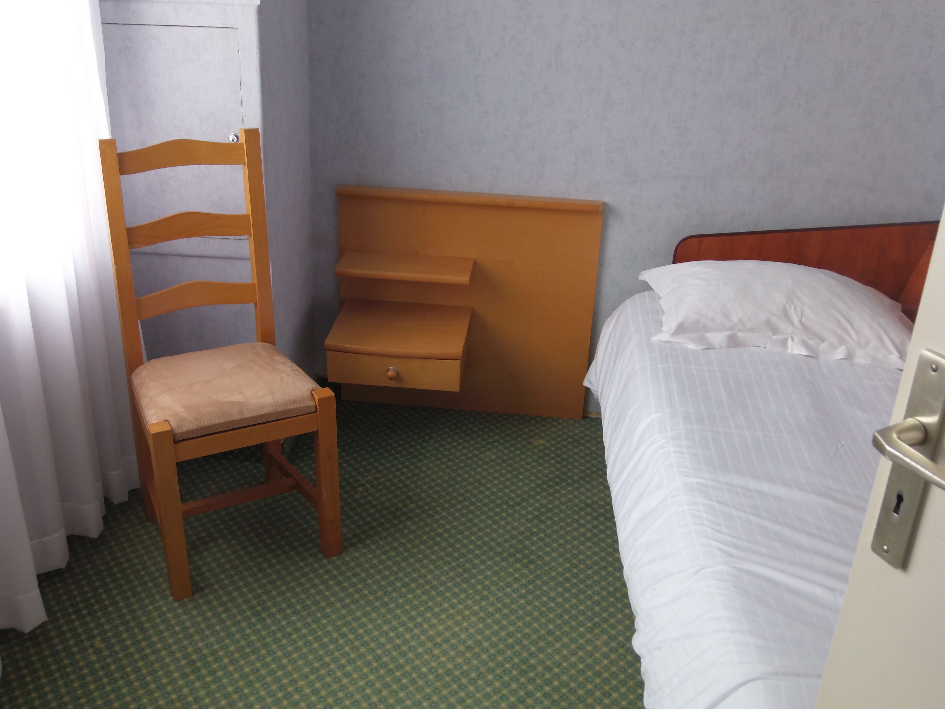 Quarto pequeno e simples com uma cama, uma cadeira de madeira e uma mesinha de cabeceira.