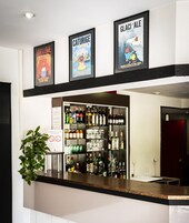 Uma área de bar com diversos frascos de bebidas e copos, um aviso proibindo fumar e três cartazes emoldurados acima do balcão.