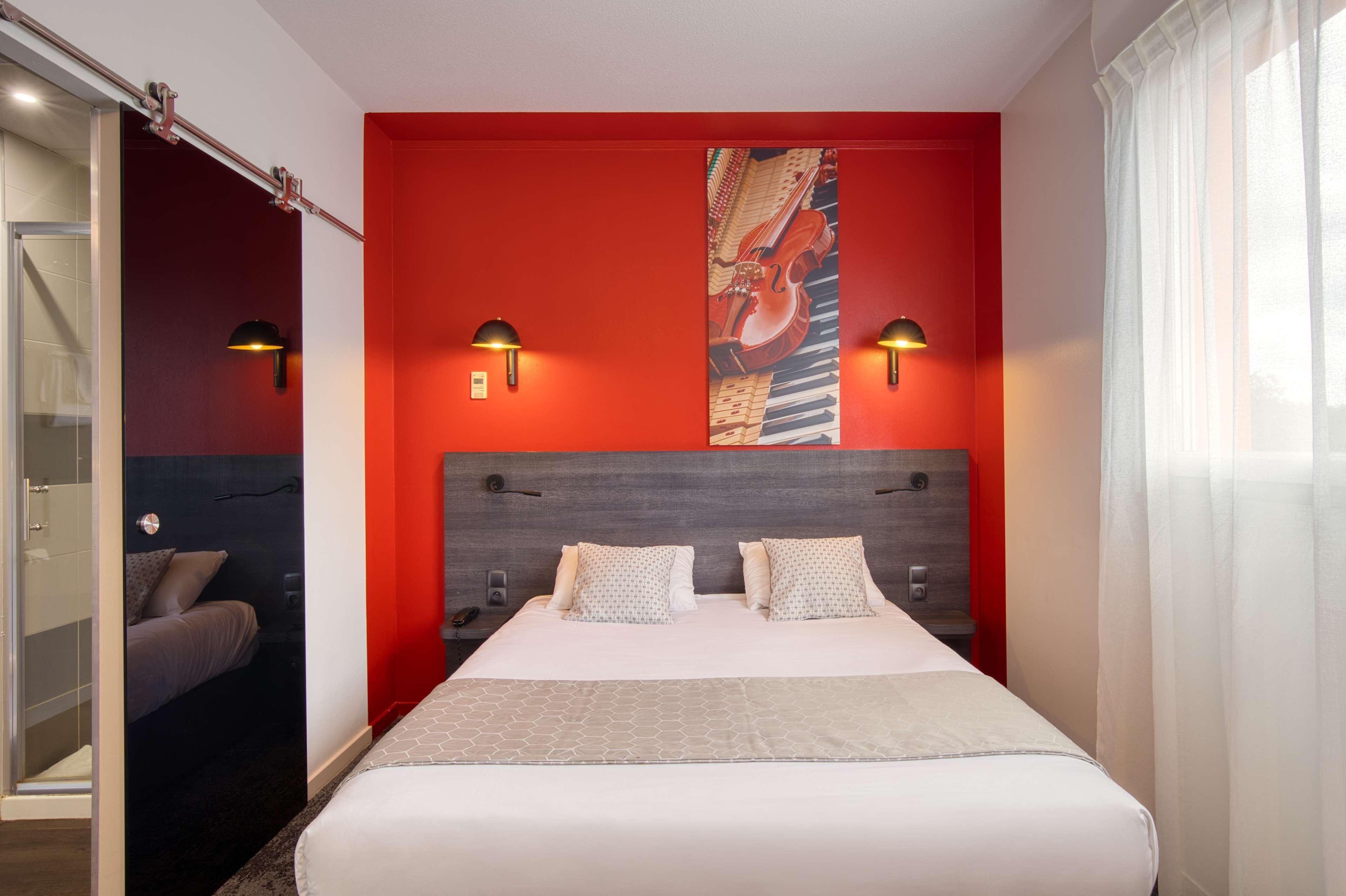 Quarto de hotel com uma cama grande, duas luminárias de cabeceira, uma parede em tom de vermelho e uma pintura de um violino.