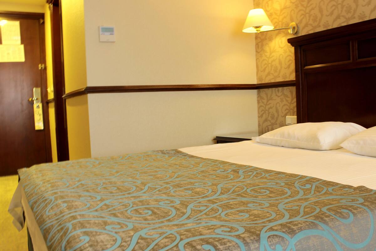 Quarto de hotel com uma cama, mesa de cabeceira, abajur e termostato na parede.