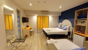 Quarto de hotel com cama, mesa de cabeceira, escrivaninha, cadeira e uma pequena cozinha.