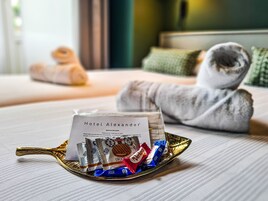 Quarto de hotel com cama, travesseiro, manta, bandeja com chocolates e um cartão.