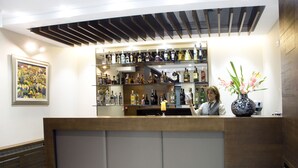 Uma área de bar com balcão de madeira, prateleiras com garrafas de bebidas e um vaso com flores.