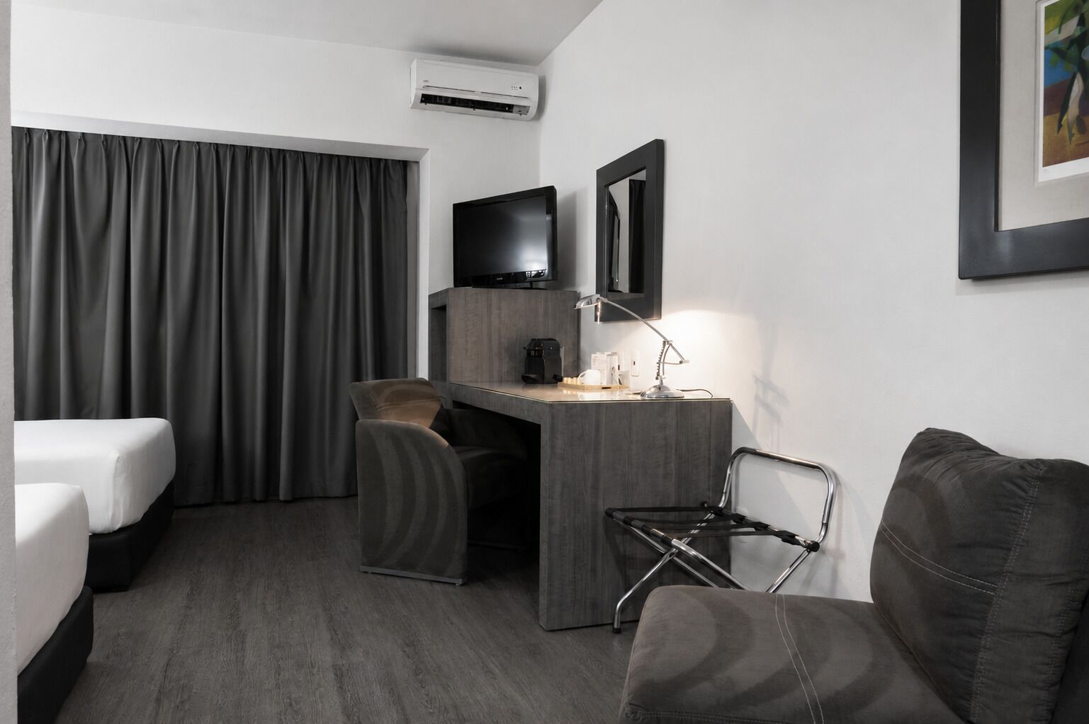 Quarto de hotel com cama, escrivaninha com cadeira, máquina de café, luminária de mesa e televisão de tela plana.
