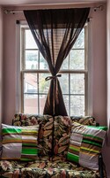 Janela com vista para um prédio e árvores, cortina amarrada no alto e uma cama com almofadas estampadas.