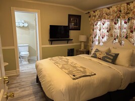 Quarto com cama, televisão, mesa de cabeceira, abajur e porta para o banheiro.
