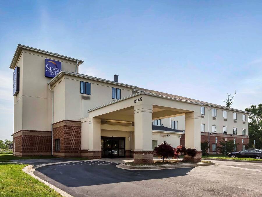 Um hotel Sleep Inn com entrada destacada, estacionamento e área ajardinada com uma pequena árvore.