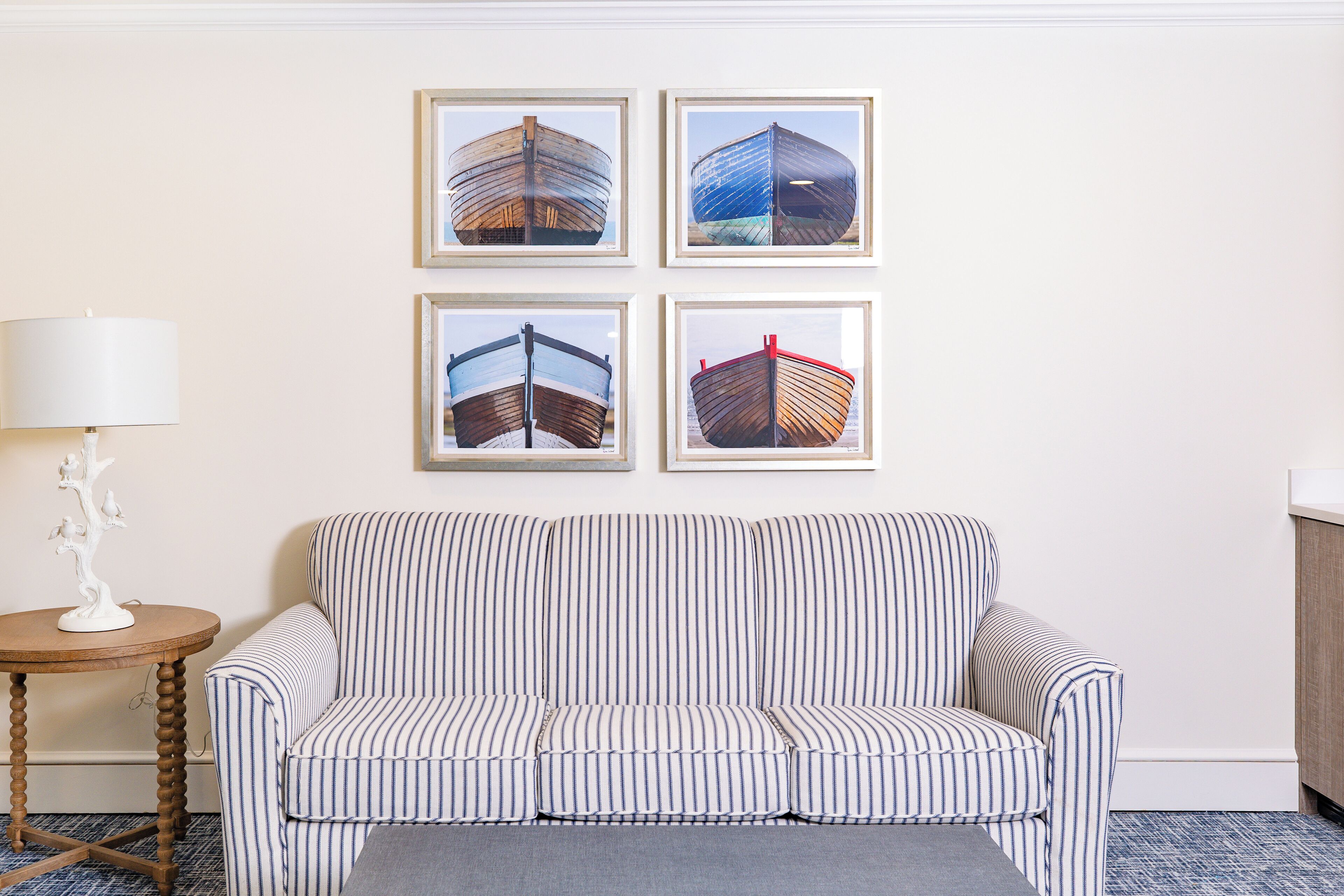 Um sofá listrado, uma mesinha redonda com uma luminária branca e quatro quadros com imagens de barcos na parede.