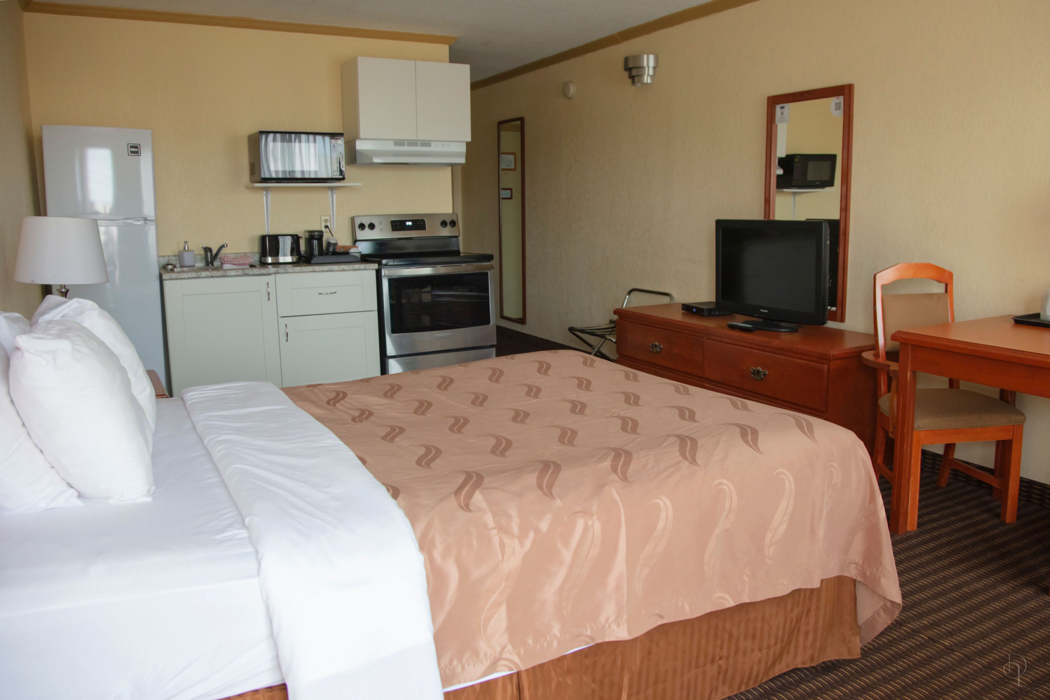 Quarto de hotel com cama, escrivaninha, televisão, cozinha compacta e espelho.