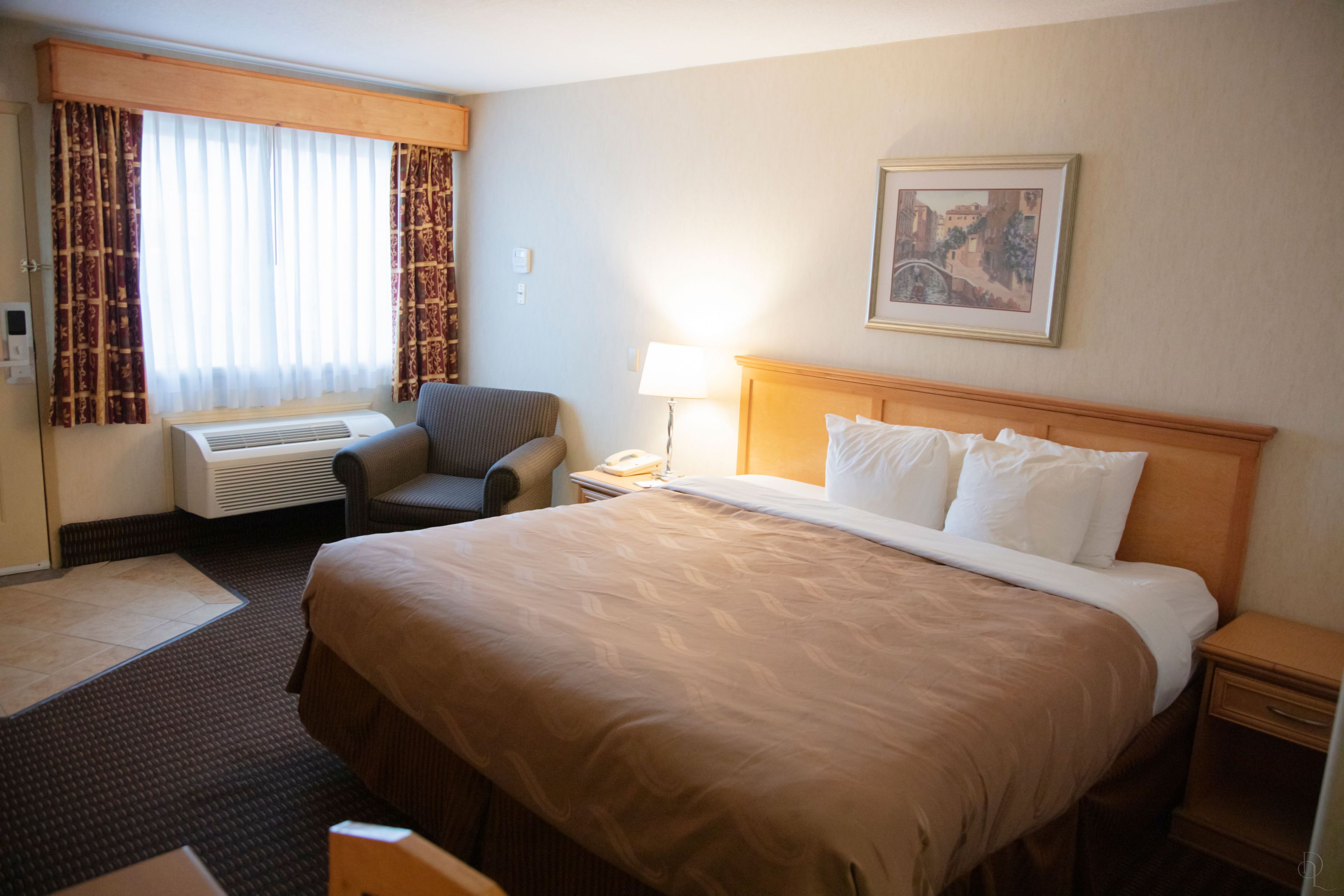 Quarto de hotel com cama grande, uma cadeira, escrivaninha e janela com cortinas.