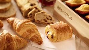 Diversos produtos de panificação, incluindo croissants e pão, dispostos numa tábua de madeira com a inscrição 'ibis kitchen'.