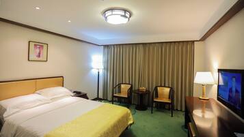 Quarto de hotel com uma cama, duas cadeiras, uma mesa com abajur, televisão e uma janela grande com cortinas.