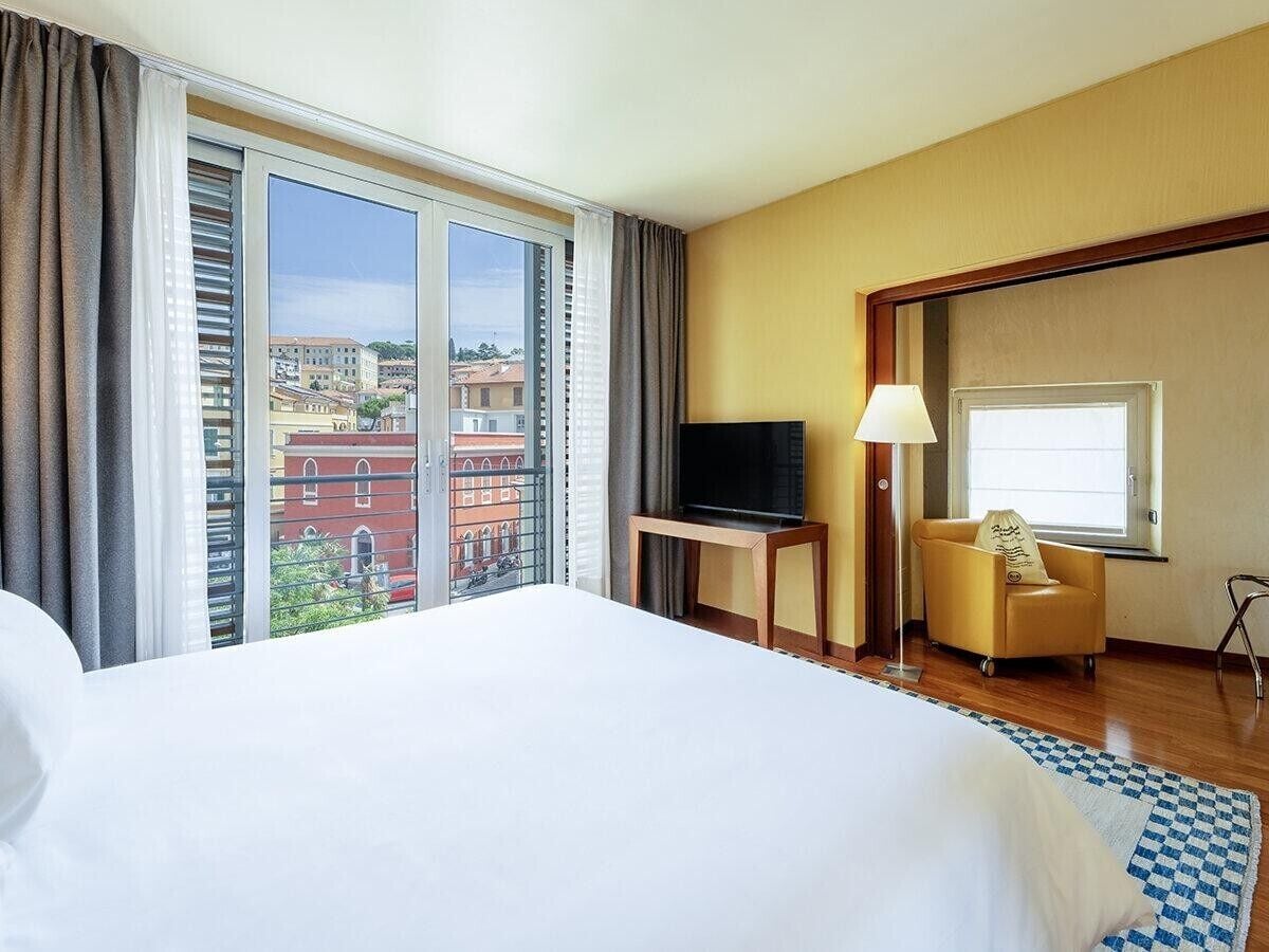 Quarto de hotel com uma cama grande, televisão, escrivaninha, cadeira e janela com vista para edifícios.
