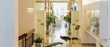 Corredor de hotel com placa de restaurante, plantas em vasos e uma vitrine de vidro.