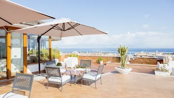 Um terraço com mobiliário de exterior, um guarda-sol grande e vista para o mar.