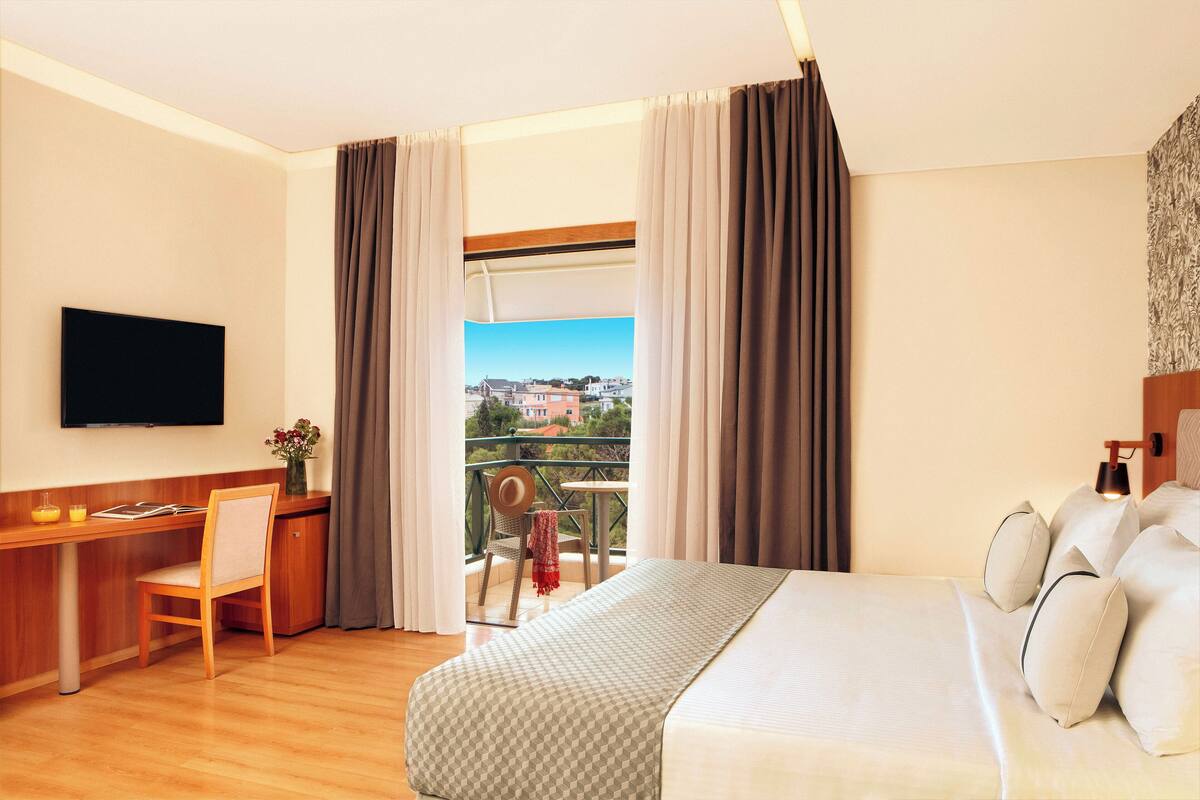 Quarto de hotel com cama, escrivaninha, cadeira, televisão e varanda com vista.