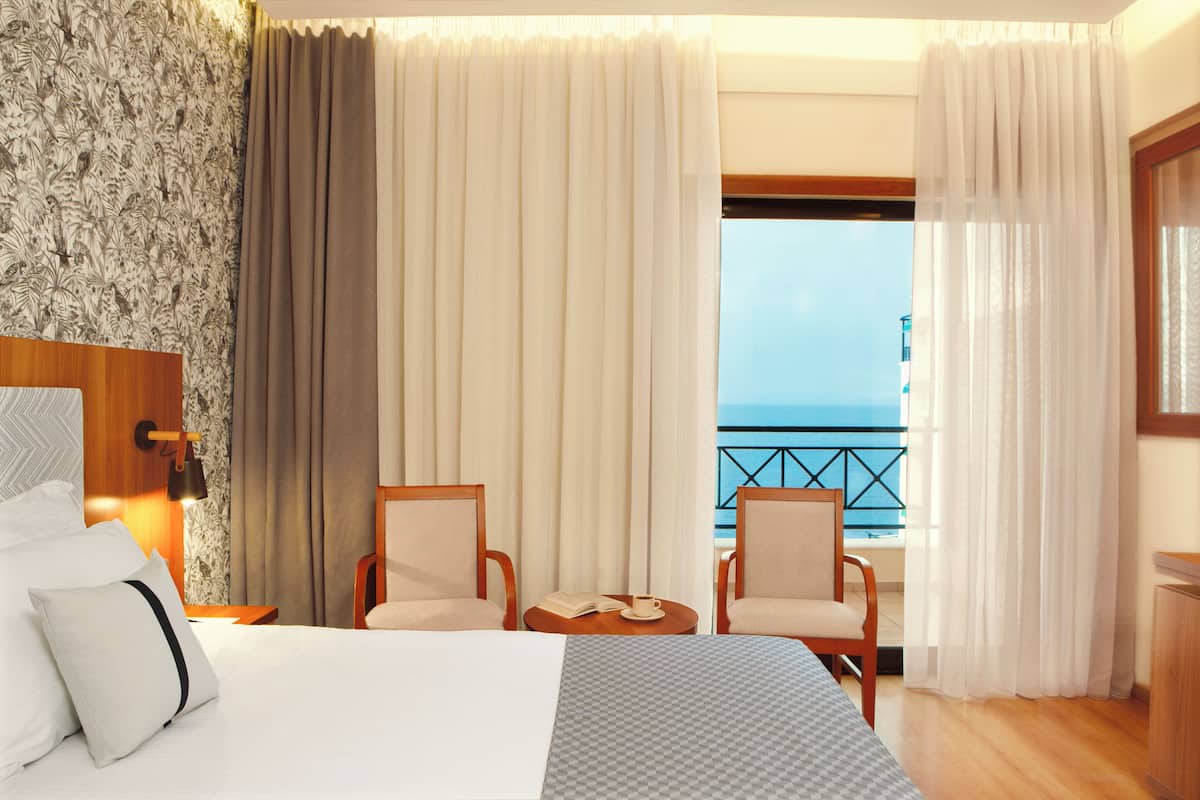 Quarto de hotel com uma cama, duas cadeiras, uma mesa pequena com um livro e uma xícara, e vista para o mar através de amplas janelas.