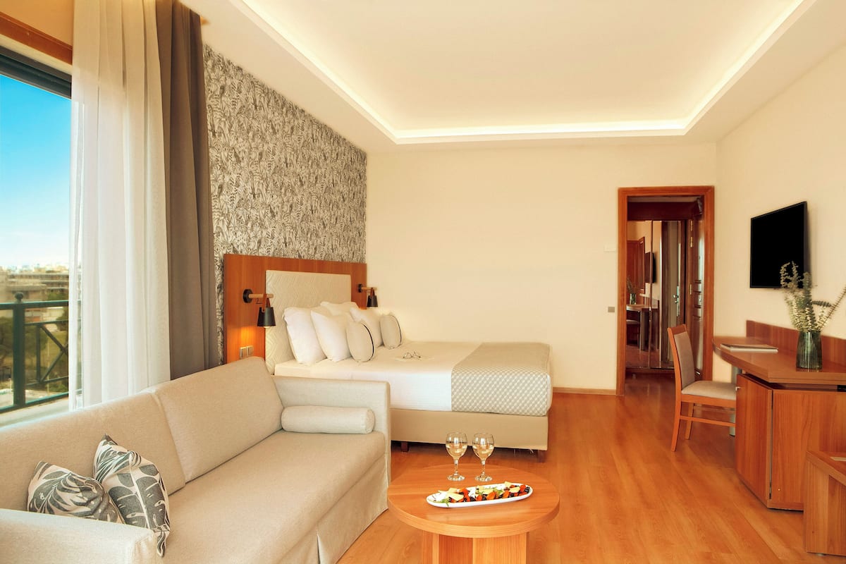 Quarto de hotel moderno com sofá, cama, mesa de jantar com prato de comida e TV fixada na parede.