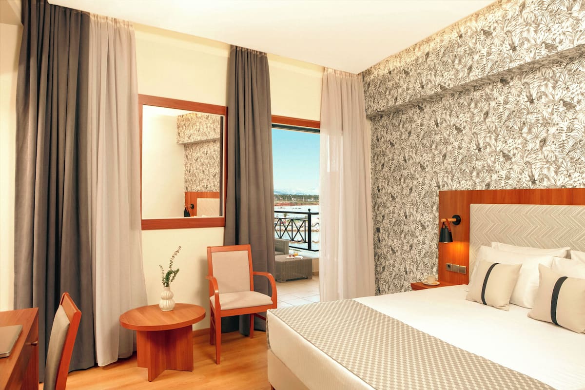 Quarto de hotel com uma cama grande, cabeceira de madeira, uma mesinha com uma cadeira, vista para o exterior e uma varanda com corrimão.