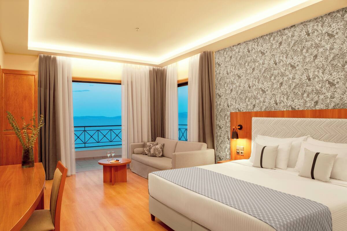 Quarto de hotel com uma cama grande, um sofá, uma mesa de jantar e vista para o mar.