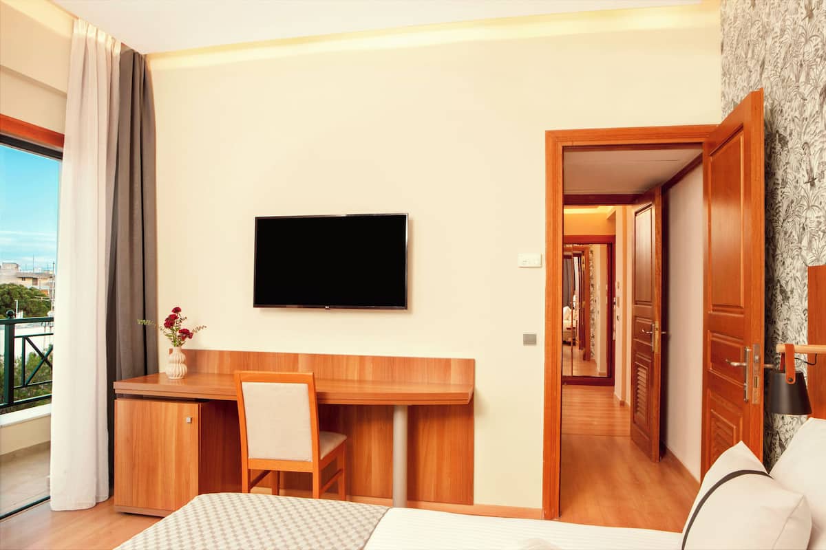 Quarto de hotel com cama, secretária com cadeira, televisão na parede e porta para um corredor.