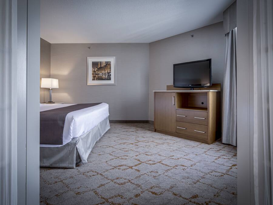 Quarto de hotel com cama, televisão em aparador de madeira, abajur e quadro na parede.