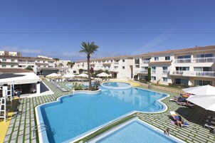 Uma área de piscina com formato singular, cercada por cadeiras de descanso e guarda-sóis, adjacente a um edifício com sacadas.