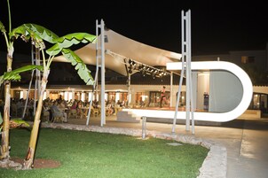 Uma área de refeições ao ar livre com um espaço coberto para sentar e um edifício moderno ao fundo.
