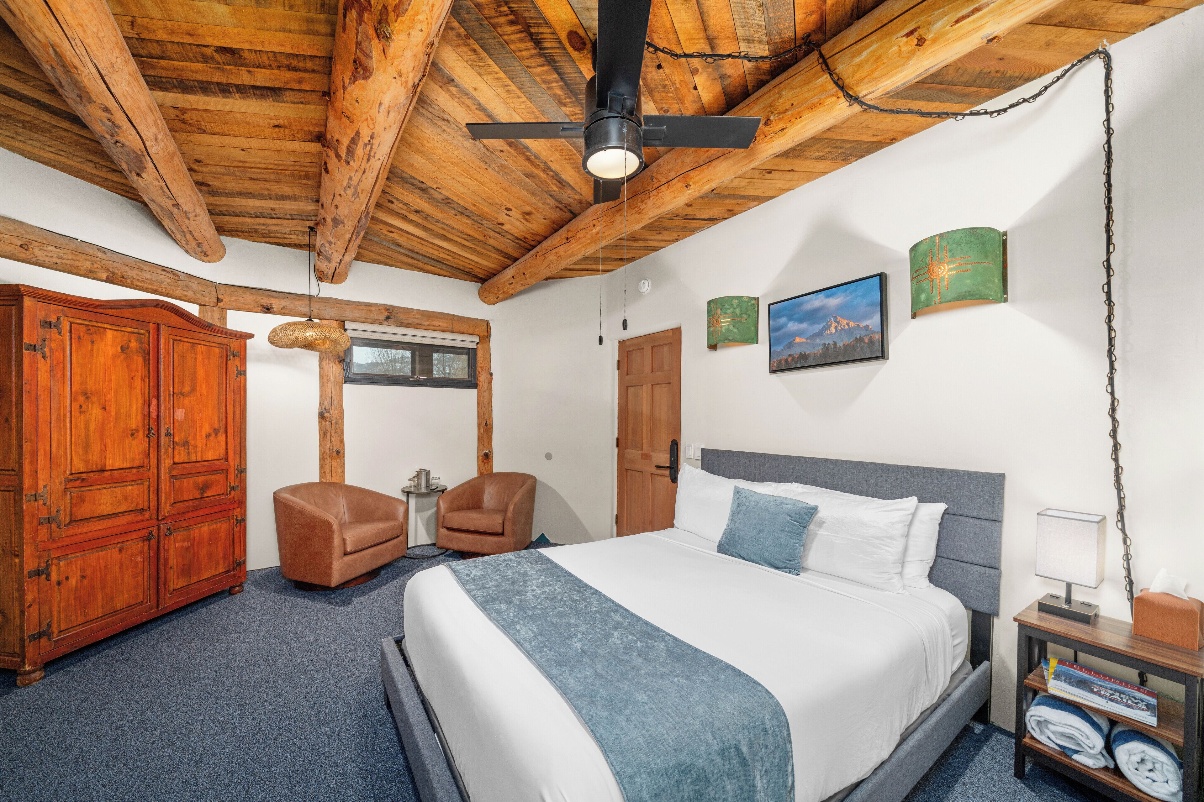 Quarto com teto de madeira, cama com roupa de cama branca e azul, guarda-roupa de madeira, poltrona de couro e uma pequena mesa com abajur e um livro.