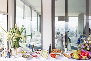 Mesa de buffet com diversos alimentos e uma garrafa de vinho, em área de jantar moderna com grandes janelas.