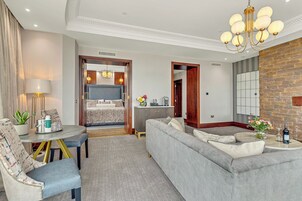 Quarto de hotel moderno com um sofá cinzento, uma mesa de jantar com cadeiras, um candelabro e vista para o quarto através de uma porta aberta.