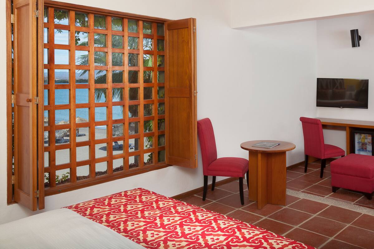 Quarto com cama, televisão, escrivaninha com duas cadeiras e uma janela grande com vista para a praia.