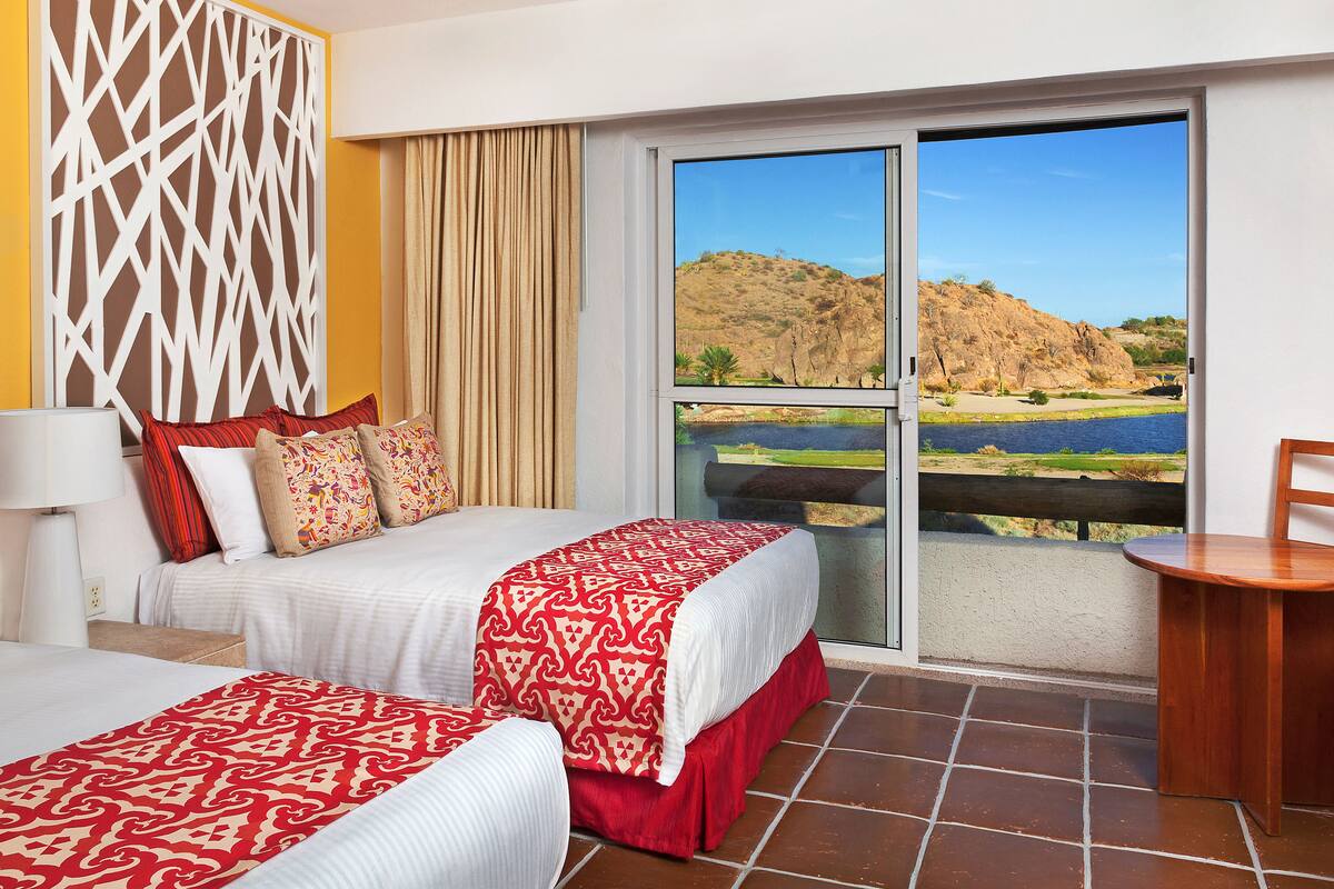 Quarto de hotel com uma cama, uma cadeira, uma mesa e vista para um rio e colinas.