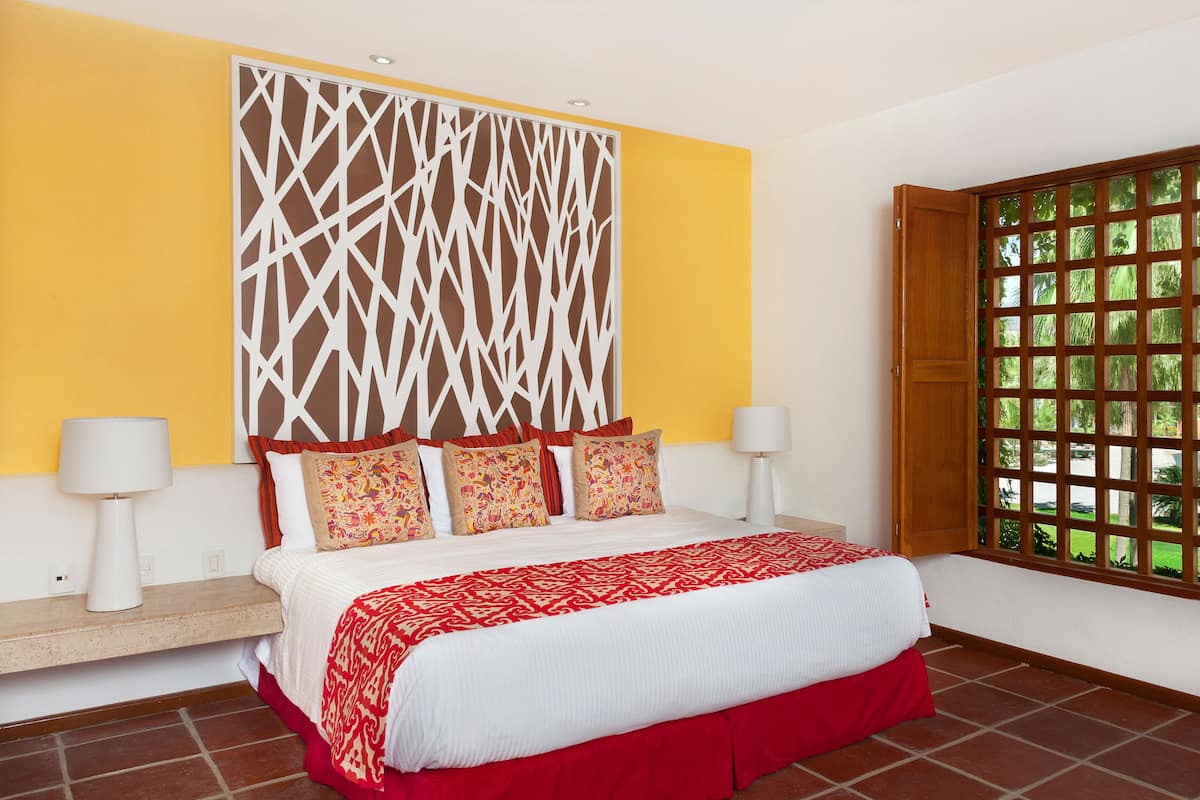 Quarto de hotel com cama, duas luminárias de cabeceira, painel decorativo na cabeceira, janela com vista e piso ladrilhado.