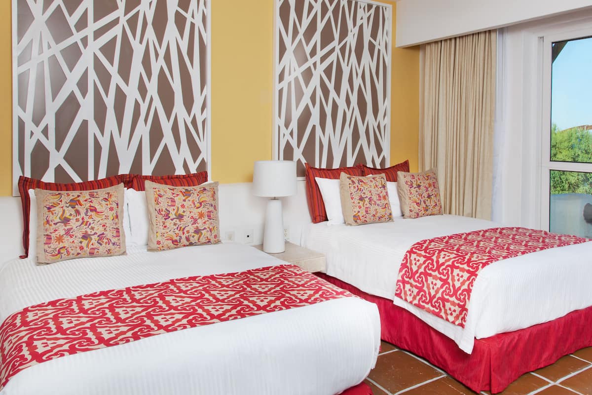 Quarto de hotel com duas camas, uma luminária branca, almofadas decorativas e vista para áreas verdes pela janela.