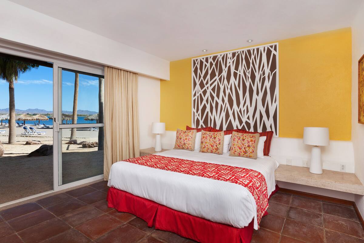 Quarto de hotel com uma cama grande, duas luminárias de cabeceira, vista para a praia e um painel decorativo na parede.