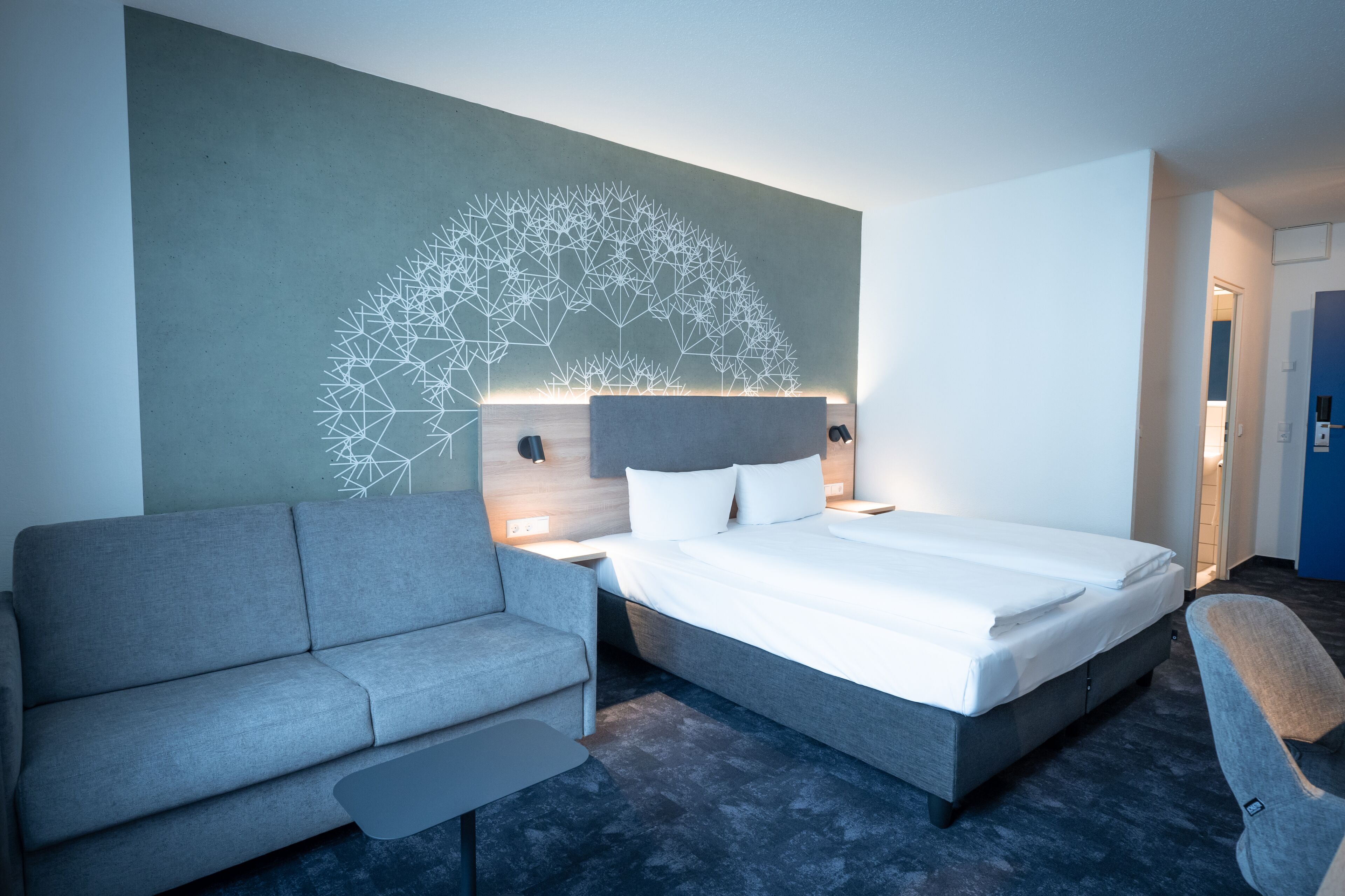 Quarto de hotel moderno com uma cama, um sofá cinza e uma obra de arte geométrica única na parede.