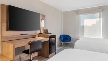 Quarto de hotel com cama, escrivaninha com cadeira, televisão e janela.