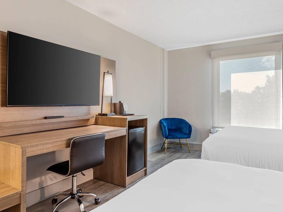 Quarto de hotel com cama, escrivaninha com cadeira, televisão e janela.
