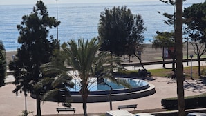 Vista de uma piscina rodeada por palmeiras e bancos, com uma praia e oceano ao fundo.