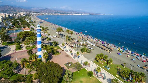 Paisagem costeira com um farol imponente, praia e edifícios urbanos.