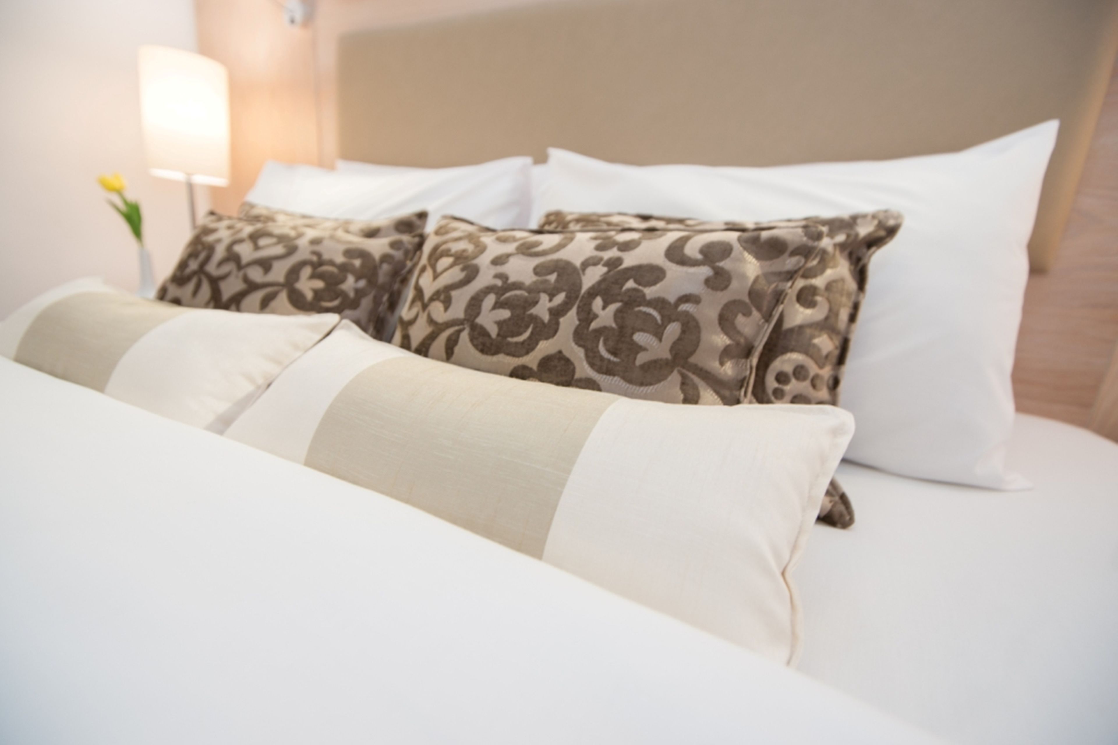 Cama bem arrumada com almofadas estampadas e lisas, abajur e um vaso com uma flor amarela.