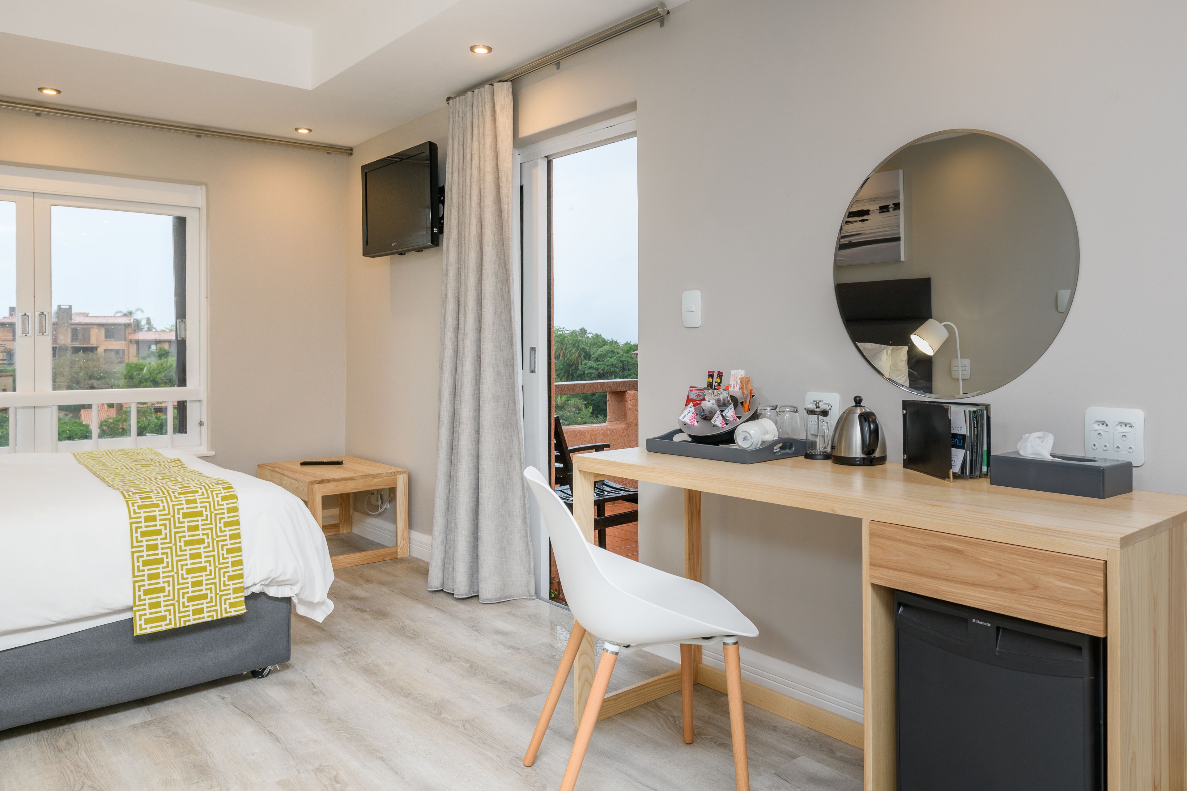 Quarto de hotel moderno com cama, escrivaninha com computador, cadeira, espelho e vista para a cidade pela janela.