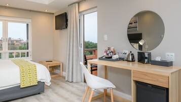 Quarto de hotel moderno com cama, escrivaninha com computador, cadeira, espelho e vista para a cidade pela janela.