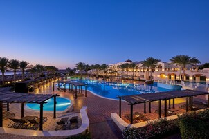 Uma grande piscina ao ar livre com áreas de estar circundantes e um edifício com janelas em arco.