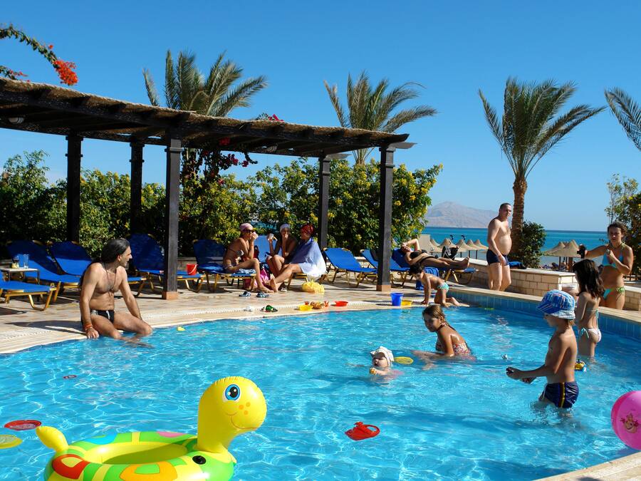 Piscina com pessoas, cadeiras de descanso e palmeiras.