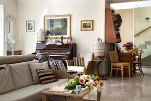 Uma sala de estar com sofá, mesa de centro e um piano. Há quadros nas paredes, uma estante de livros e uma poltrona.