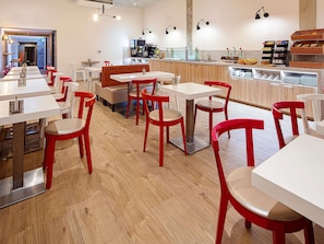 Um café moderno com piso de madeira, cadeiras vermelhas, mesas brancas e um buffet com diversas opções de comida.